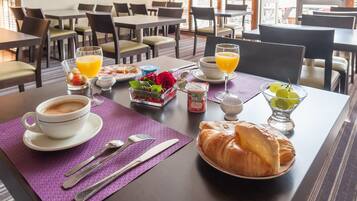 Mesa de café da manhã com um croissant, suco de laranja, café e uma cesta de frutas em uma área de refeições de hotel.