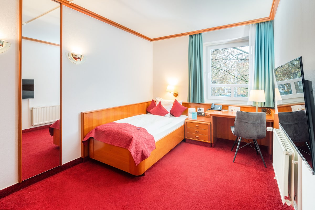 Quarto de hotel com cama, escrivaninha, cadeira, janela com cortinas, espelho e radiador.