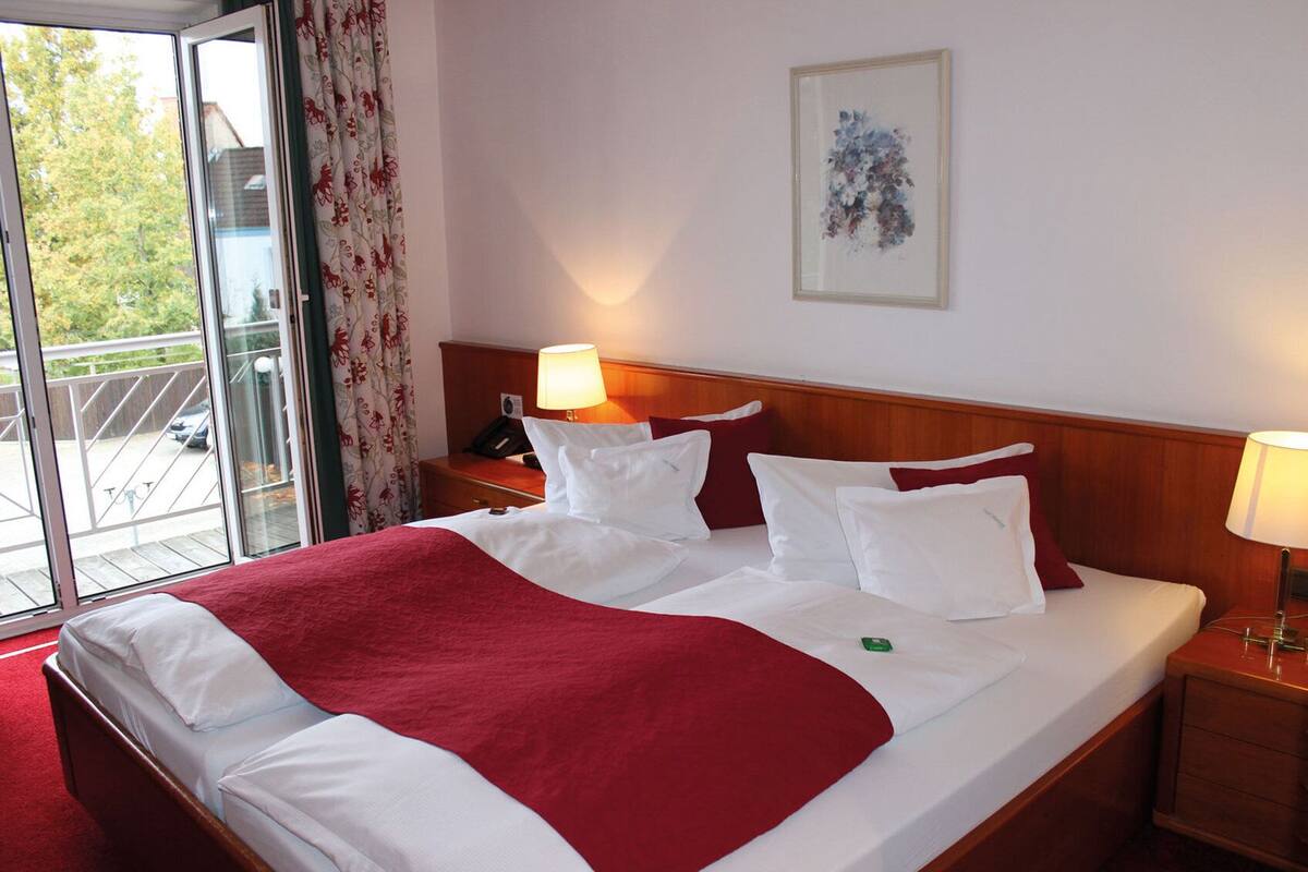 Quarto de hotel com uma cama grande, uma mesa de cabeceira com abajur, uma obra de arte emoldurada na parede e uma porta de vidro deslizante para o exterior.