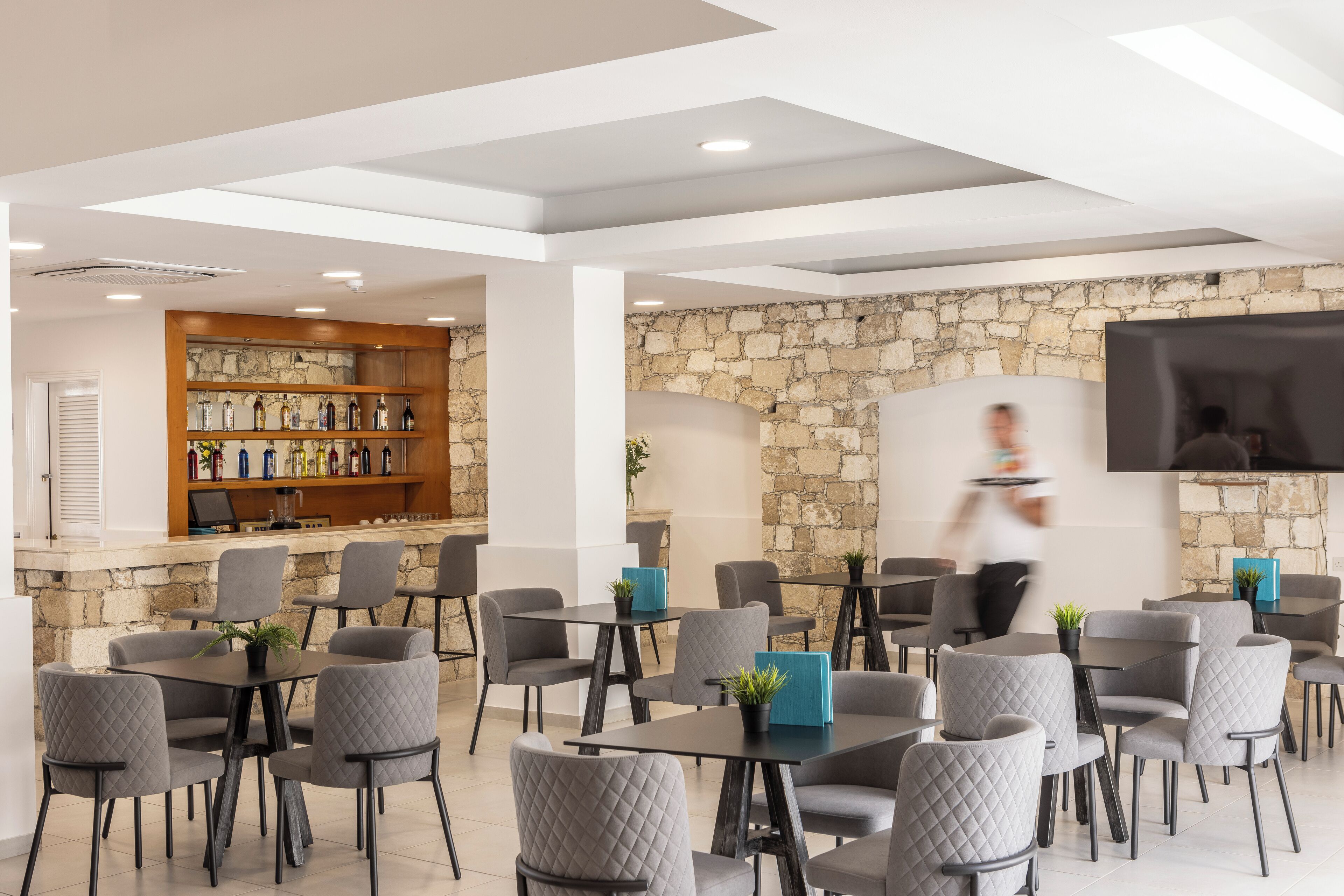 Uma área de estar moderna com várias mesas e cadeiras, uma parede de pedra, um bar com prateleiras de garrafas e uma TV de tela plana na parede.