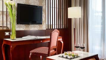Quarto de hotel com escrivaninha de madeira, televisão, cadeira, mesa com bebidas e janela com cortinas.