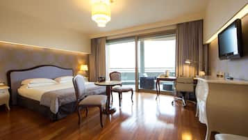 Quarto de hotel com uma cama grande, duas cadeiras, uma escrivaninha, televisão de tela plana e varanda com cortinas.