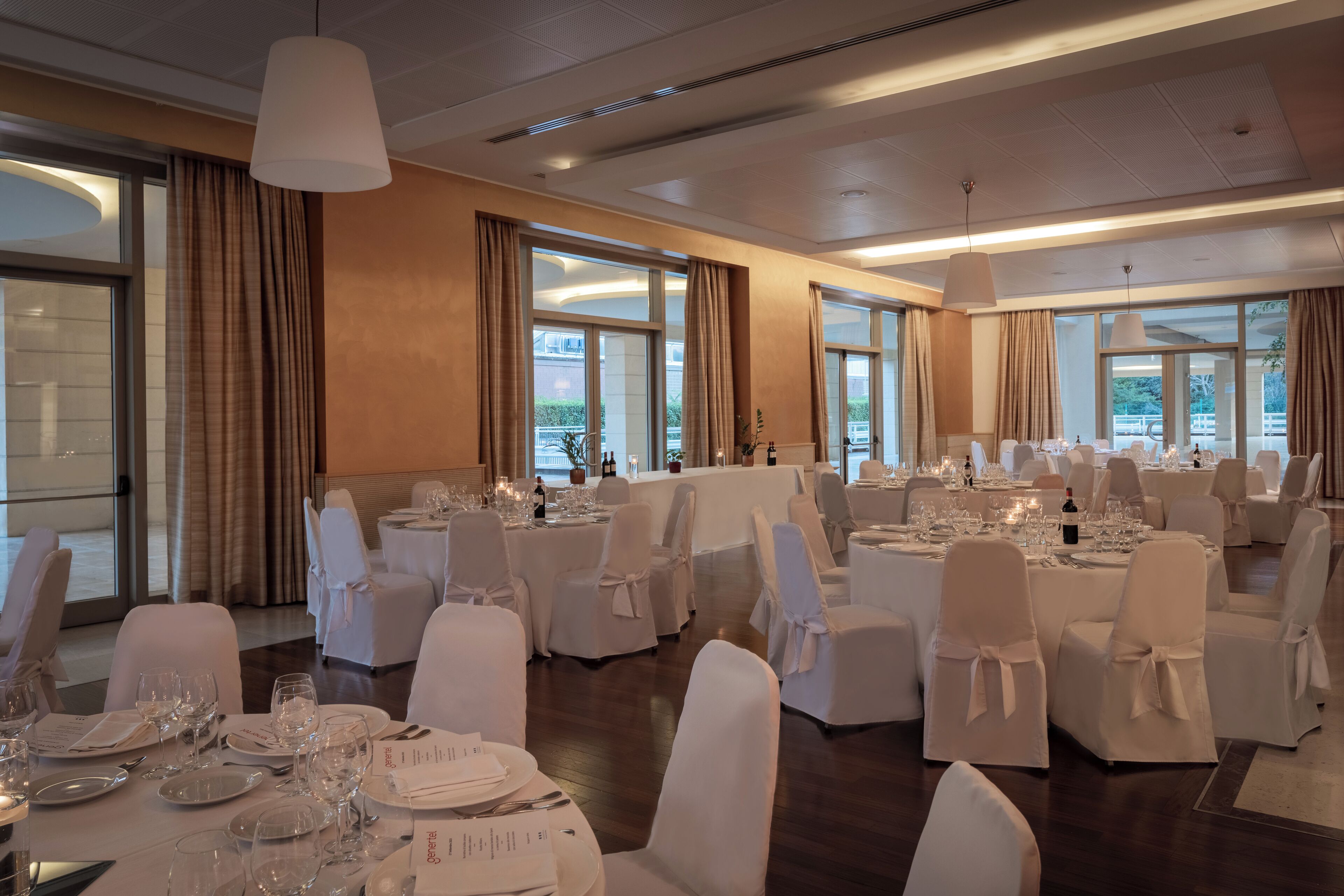 Uma sala de banquetes com mesas redondas preparadas para refeições, cadeiras brancas e amplas janelas que proporcionam iluminação natural.