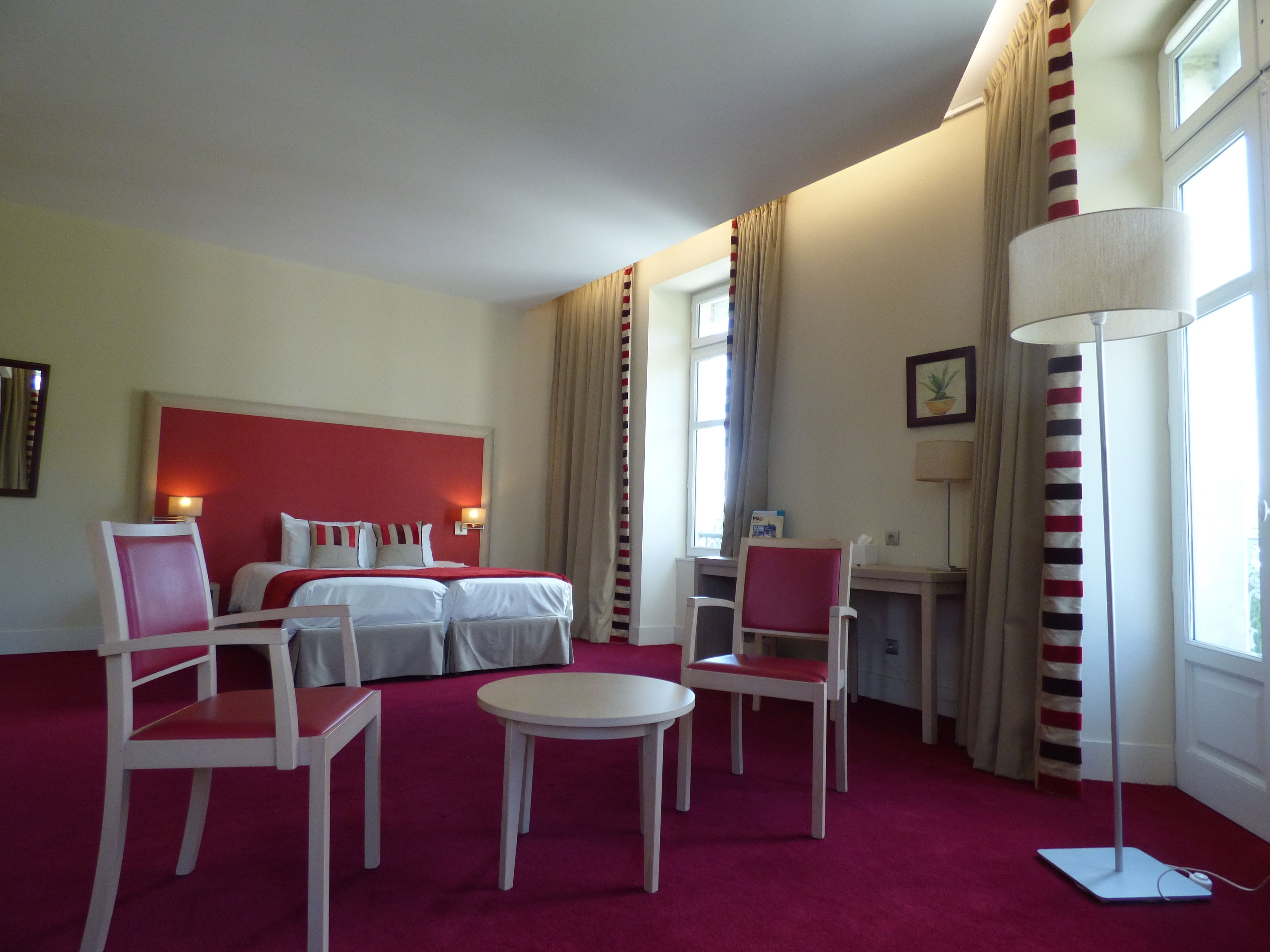 Quarto de hotel com carpete vermelho, duas camas com roupa branca, uma escrivaninha com cadeira, um abajur e uma janela com cortinas.