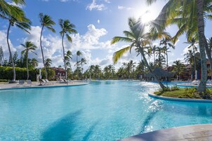 Uma piscina grande e convidativa cercada por palmeiras e espreguiçadeiras, com um bangalô de palha e um edifício ao fundo.