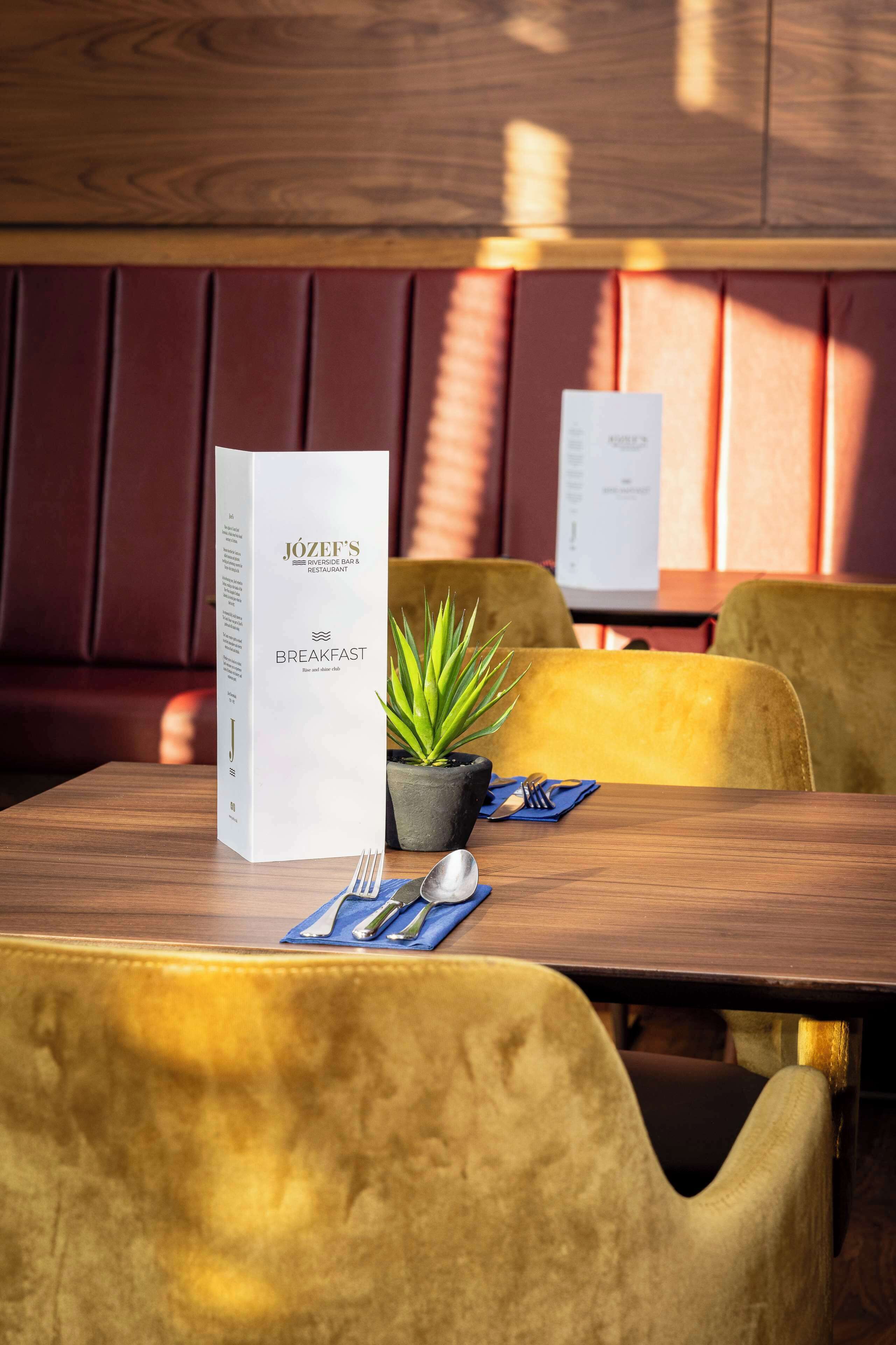Ambiente de restaurante com mesa de madeira, caixa para menus, uma planta e menu na parede.