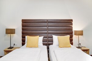Quarto de hotel com uma cabeceira grande e acolchoada, duas camas com roupa de cama branca e duas luminárias de cabeceira.