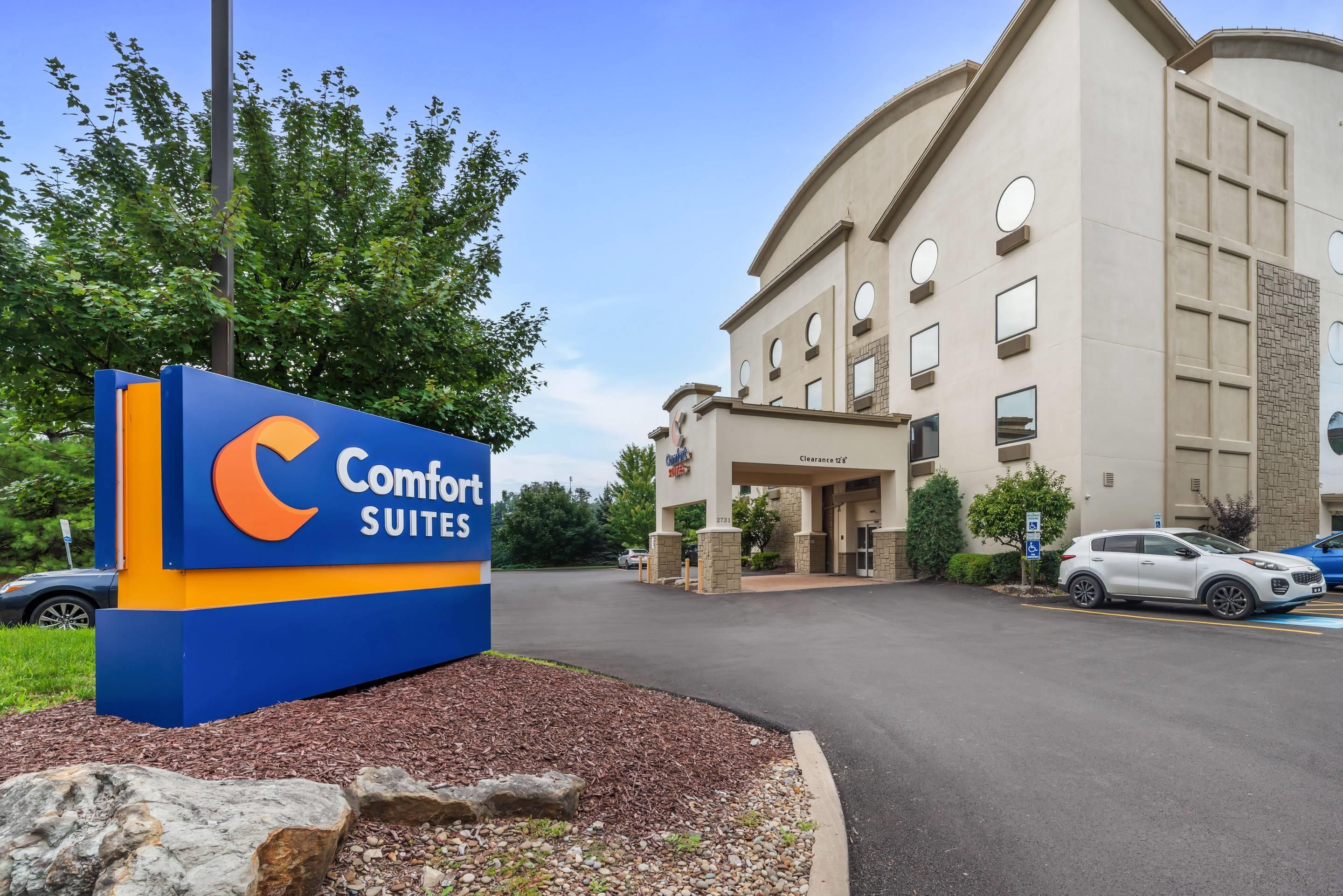 Entrada de hotel com placa da Comfort Suites, estacionamento e um edifício com várias janelas.