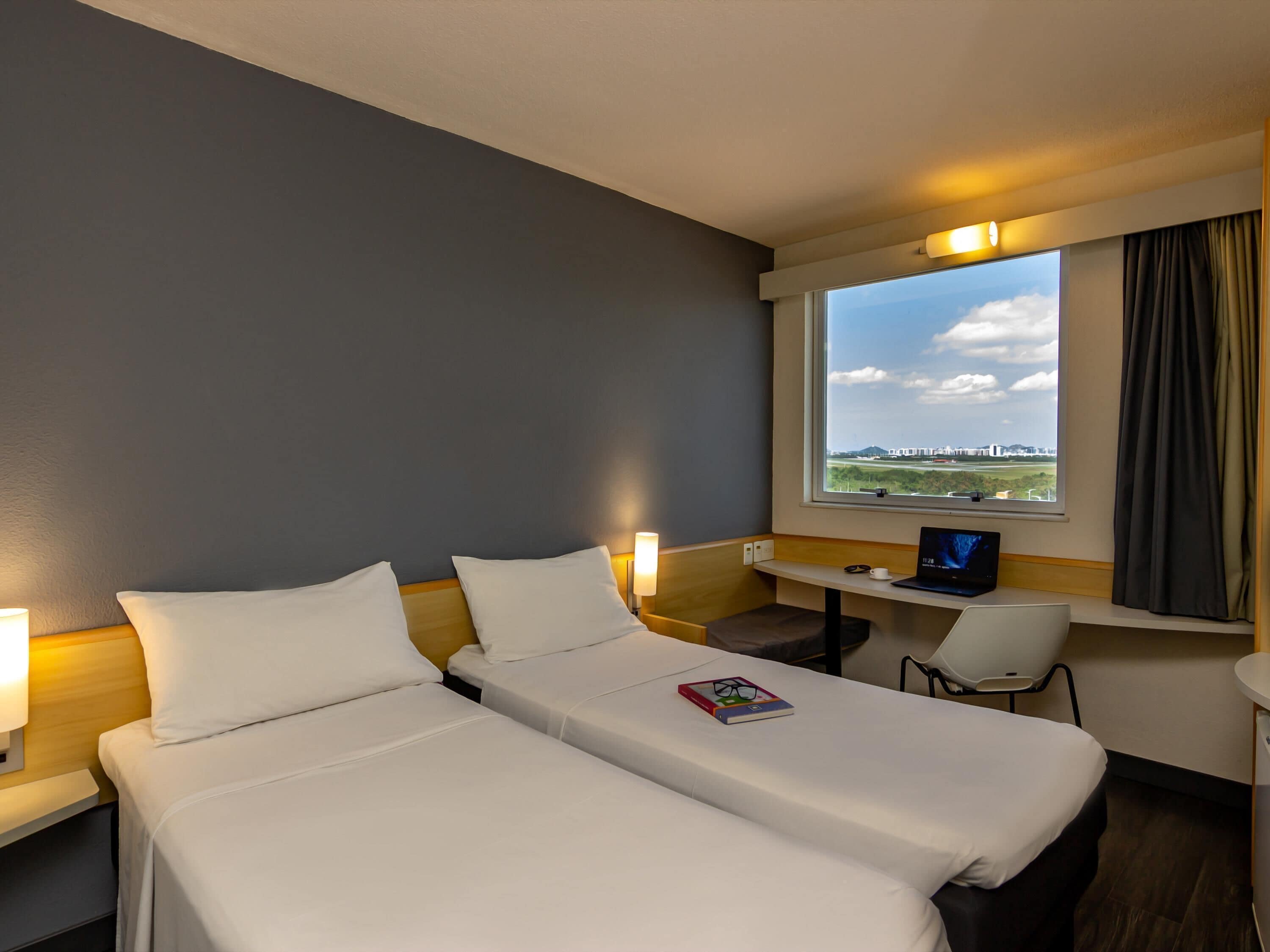 Quarto de hotel com duas camas, uma escrivaninha com um laptop, uma cadeira e uma janela com vista para a paisagem urbana.