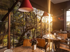 Restaurante com vista para vegetação exuberante através de grandes janelas, mobiliário de madeira e luminárias pendentes vermelhas.