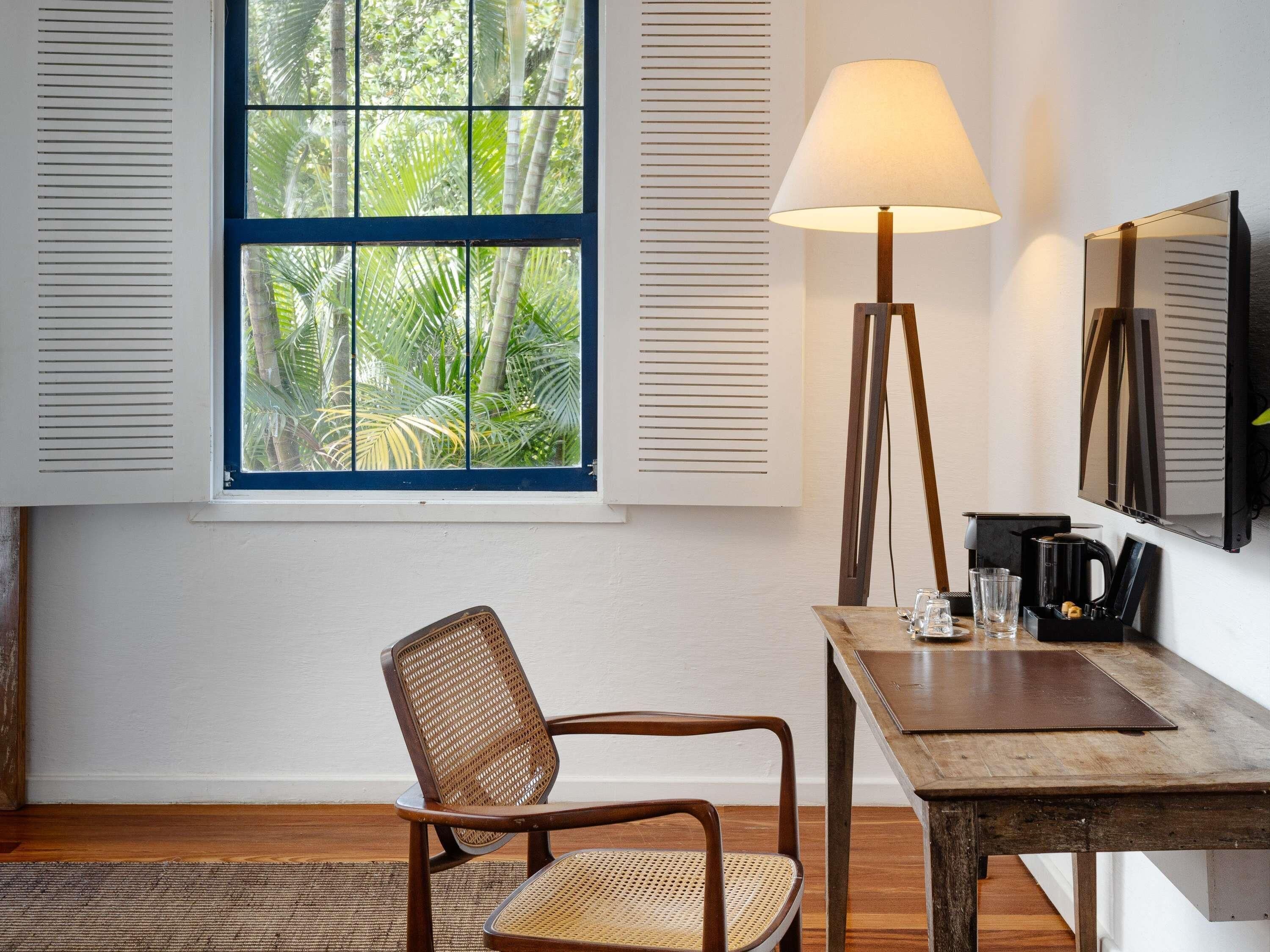 Um quarto com uma escrivaninha de madeira, uma cadeira, uma luminária, uma cafeteira e uma janela com persianas.
