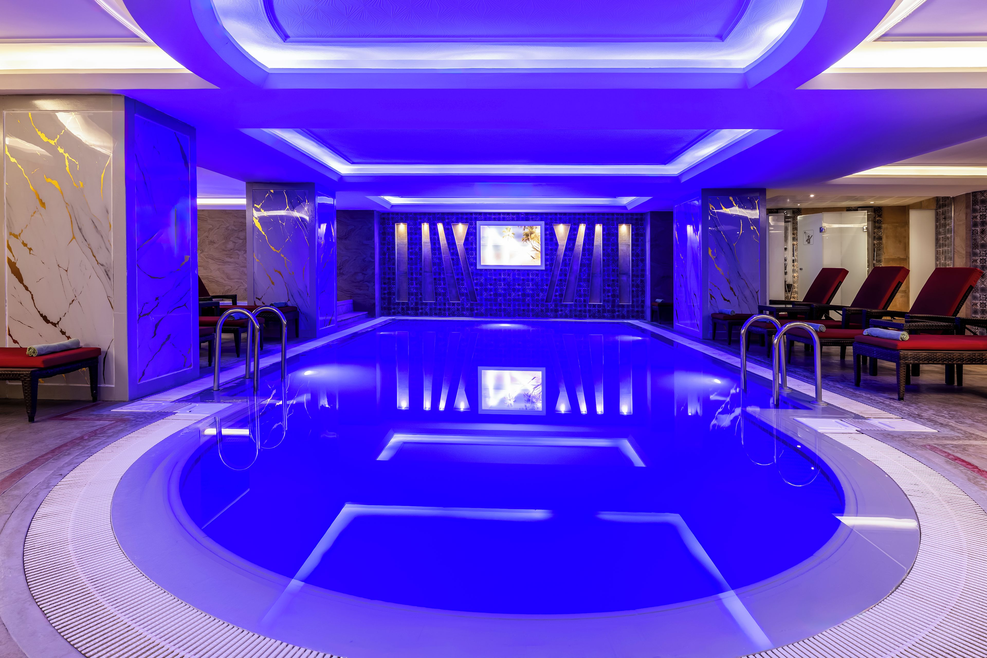 Piscina interior moderna iluminada em azul, com uma tela grande e cadeiras de descanso vermelhas.