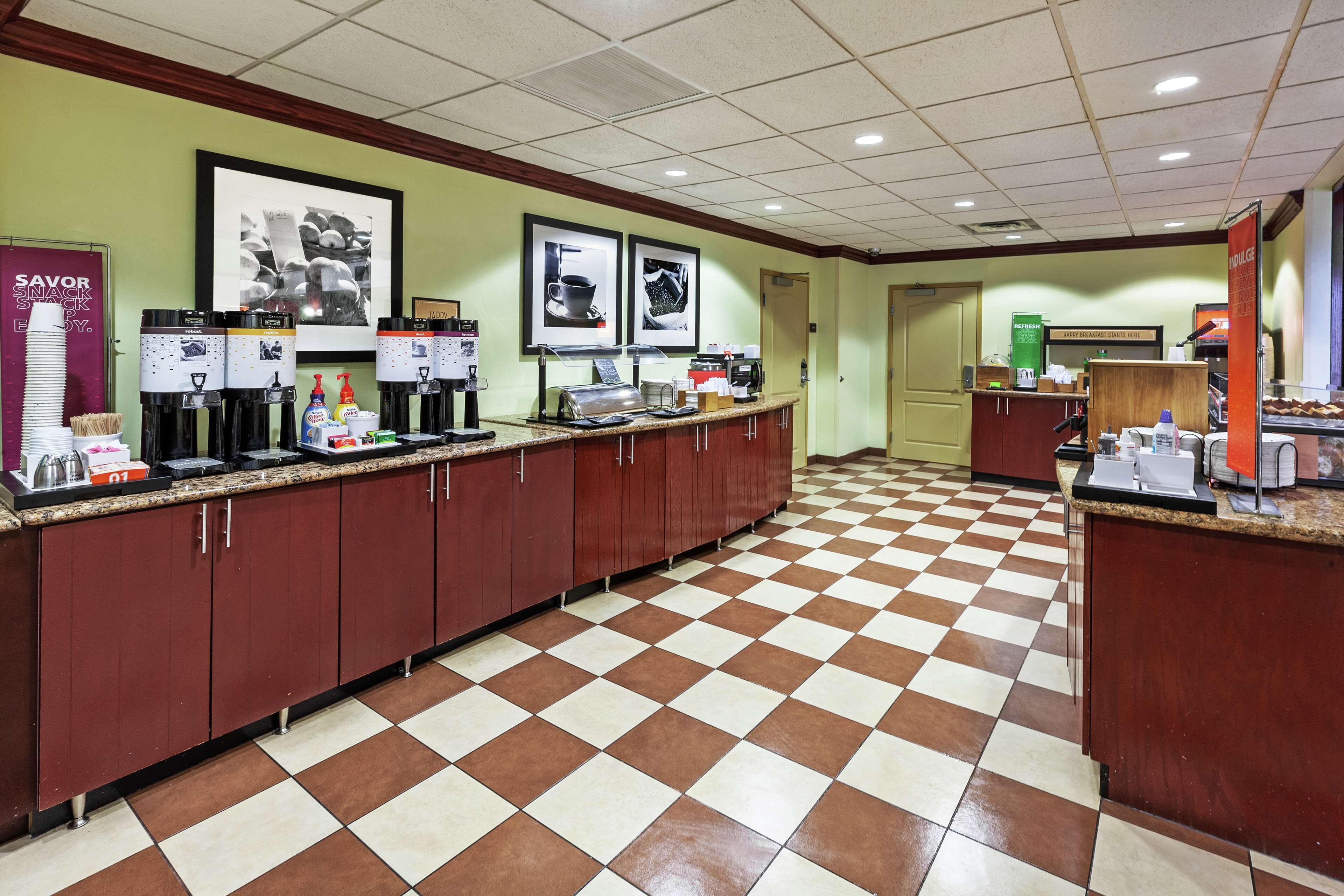 Cozinha de hotel com chão xadrez, um espaço para café e um aviso indicando um bar de petiscos.