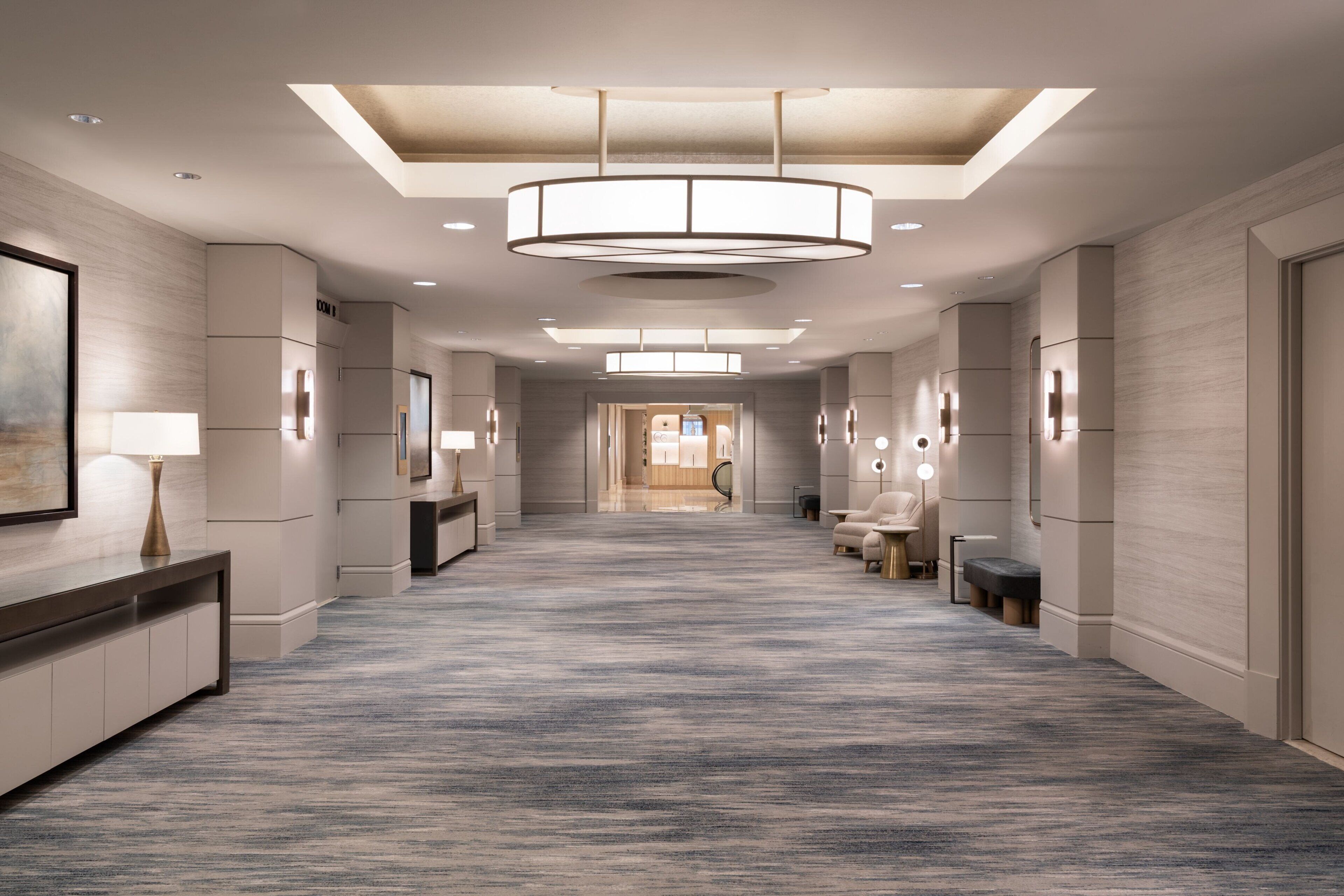 Corredor amplo de hotel com carpete estampado, iluminação embutida e um lustre central.