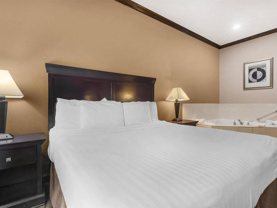 Quarto de hotel com uma cama grande, duas luminárias de cabeceira, mesa de cabeceira, banheiro com banheira e uma moldura com uma imagem na parede.