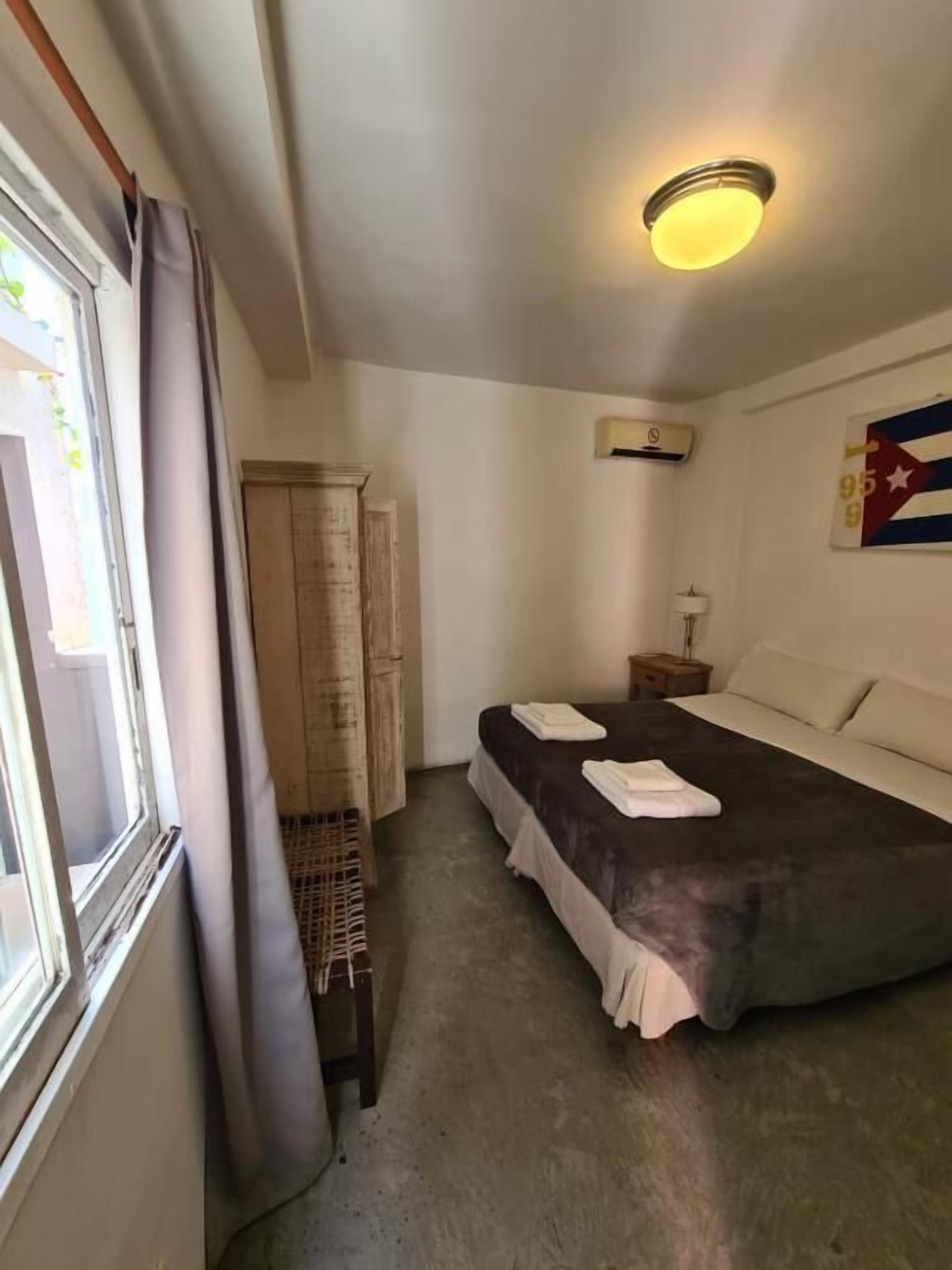 Quarto de hotel com cama, guarda-roupa, mesa pequena e ar condicionado fixado na parede.