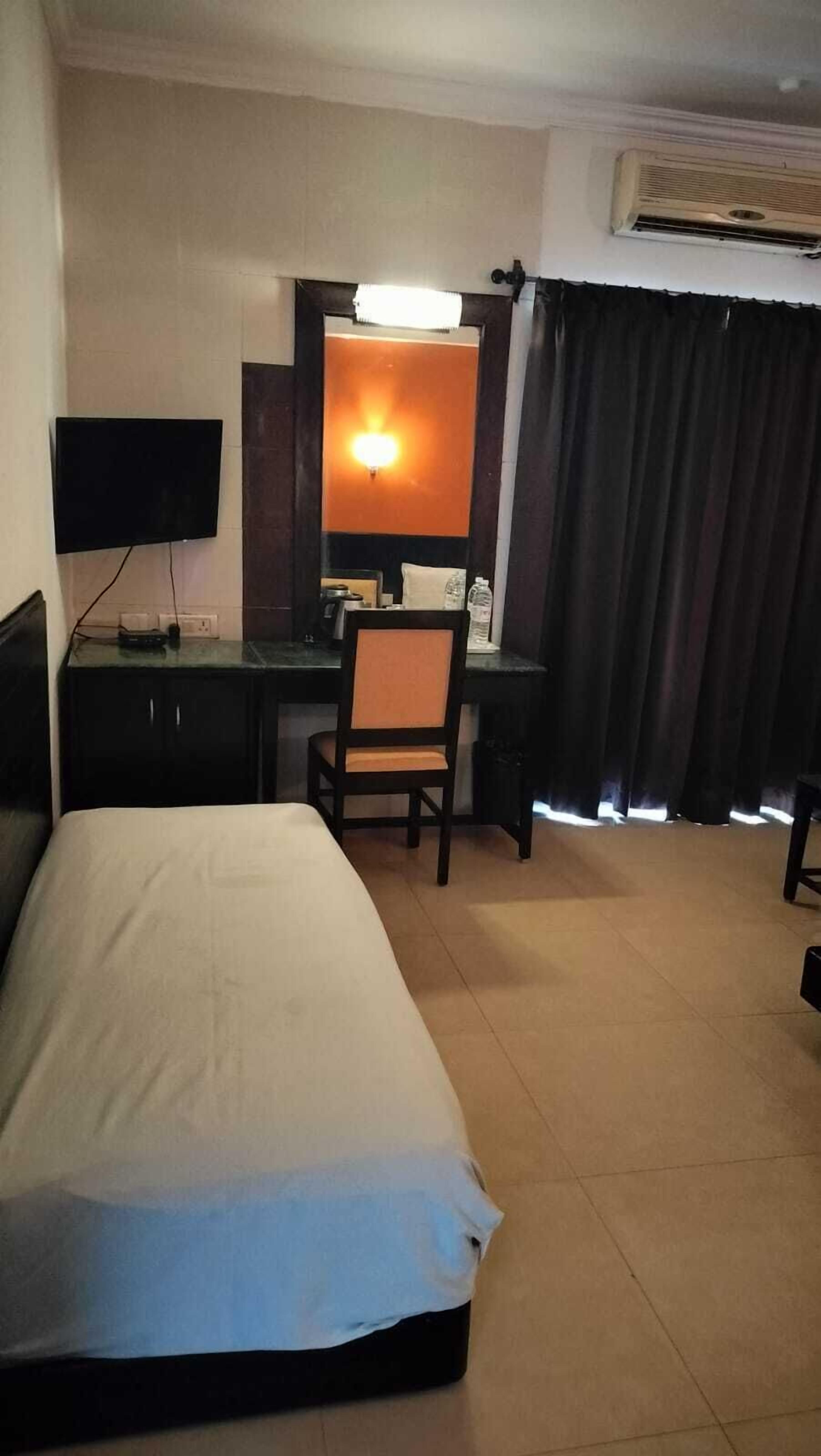 Quarto de hotel com cama, escrivaninha, cadeira, televisão e janela com cortinas.