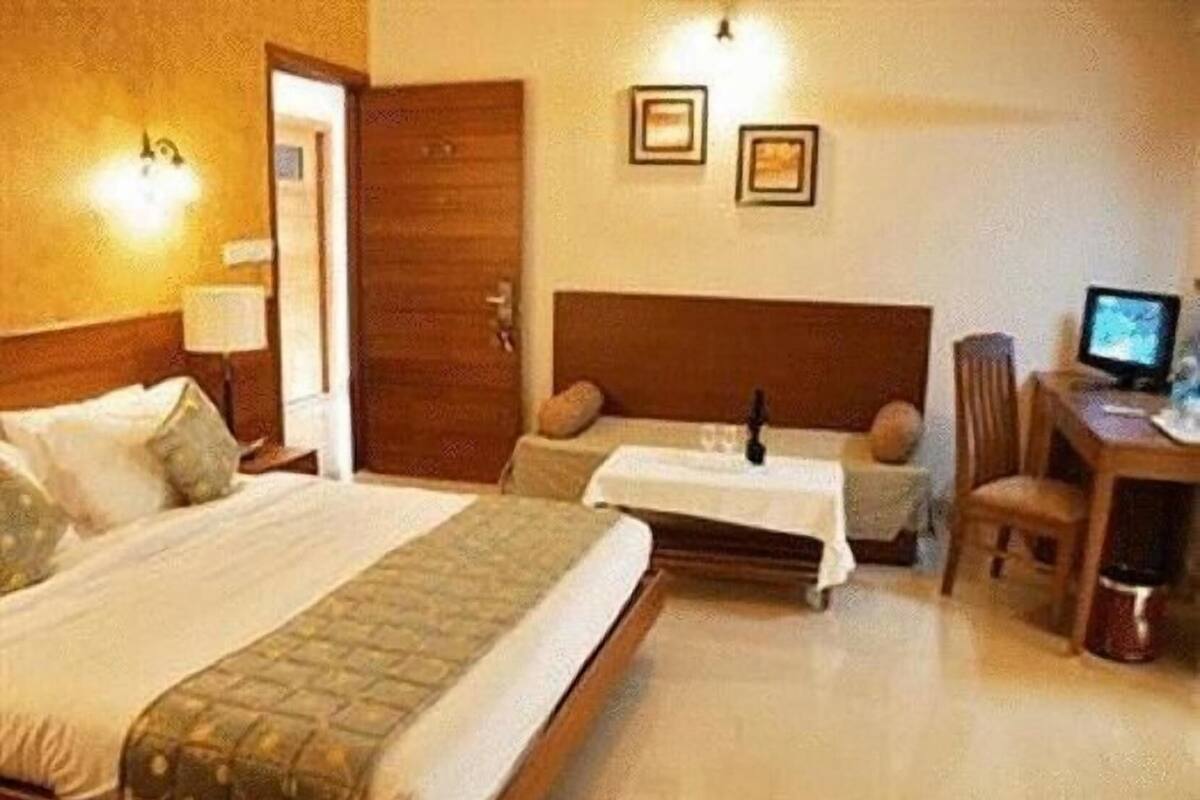 Quarto de hotel com cama, mesa de cabeceira, abajur, cadeira, escrivaninha, computador e duas imagens emolduradas na parede.