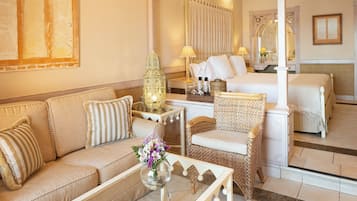 Quarto de hotel com sofá, uma pequena mesa com um vaso de flores, uma cadeira de vime, cama com roupa de cama branca e um espelho grande.