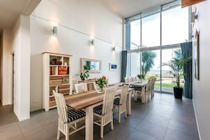 Uma sala de jantar ampla com uma grande mesa de madeira e cadeiras brancas, uma estante embutida e vista para o exterior através de amplas janelas.