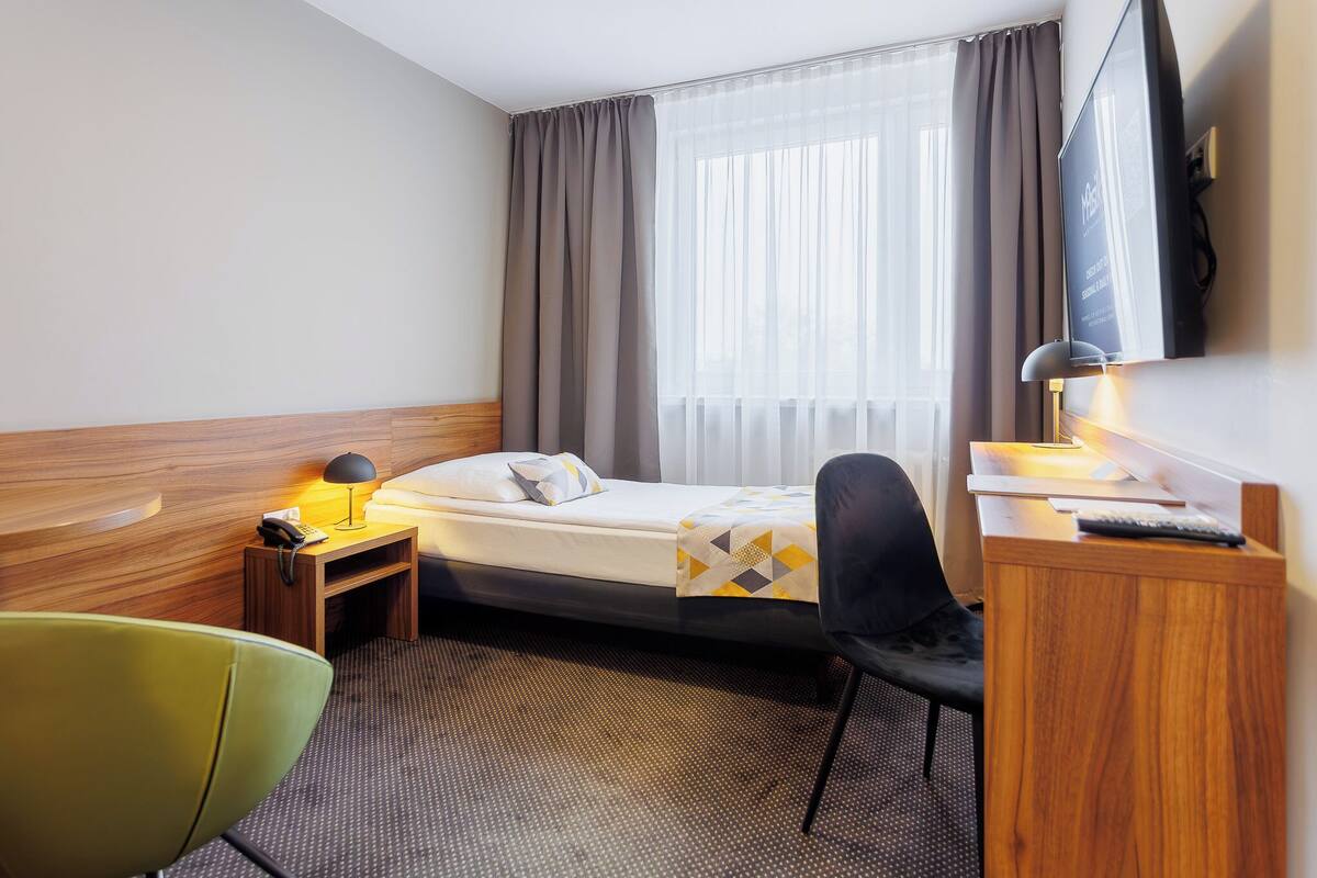 Quarto de hotel com cama, escrivaninha com luminária, cadeira, televisão e janela com cortinas.