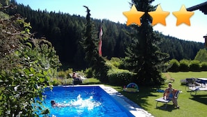 Uma piscina rodeada por um terraço de pedra, com uma área gramada e árvores ao fundo.
