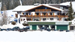Um edifício de vários andares com um teleférico ao fundo, rodeado por árvores e montanhas cobertas de neve.