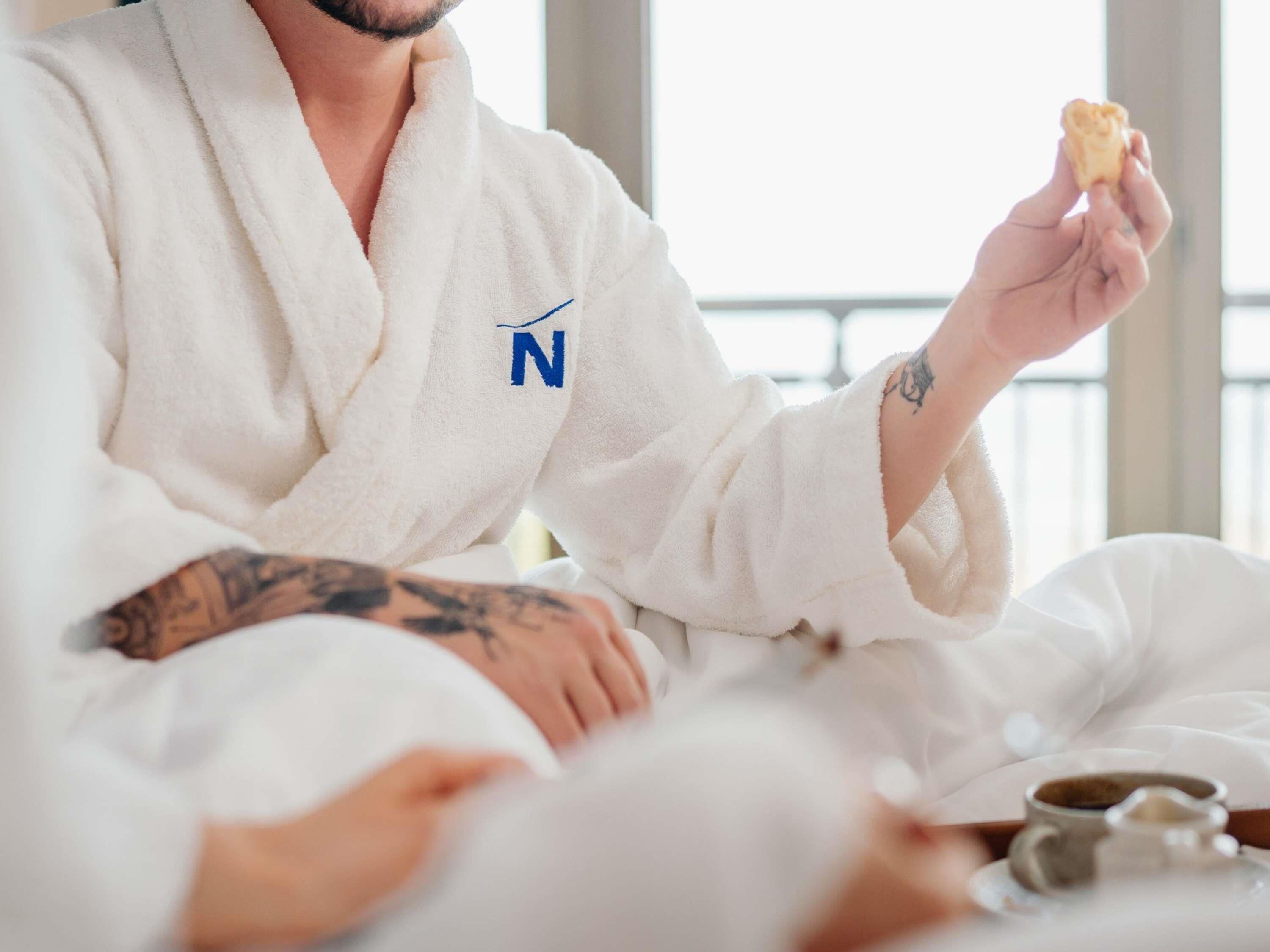 Uma pessoa vestindo um robe branco, com uma tatuagem visível no braço, sentada em um quarto com uma janela e uma xícara sobre a mesa.