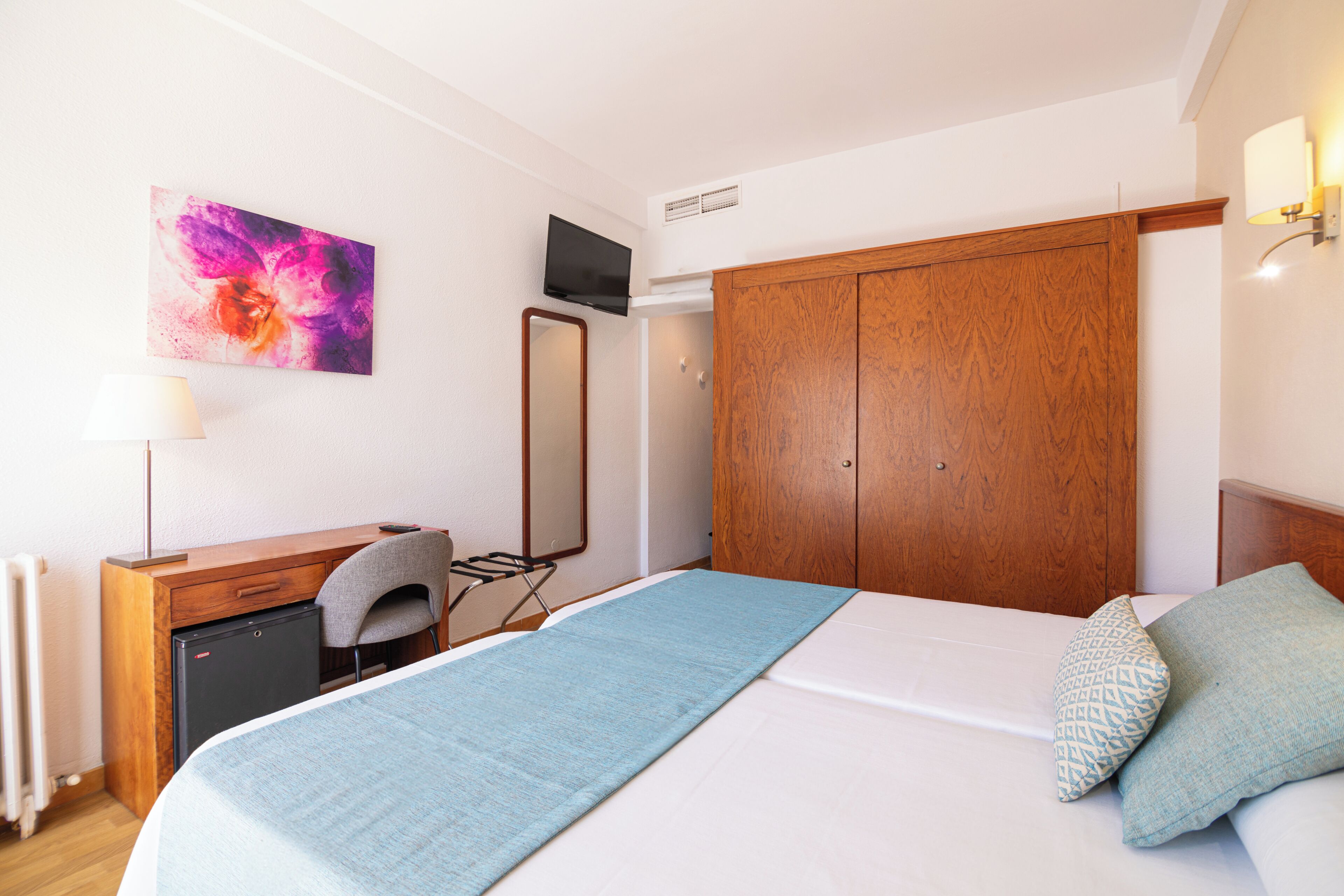 Quarto com cama, escrivaninha, cadeira, guarda-roupa, televisão e um quadro.