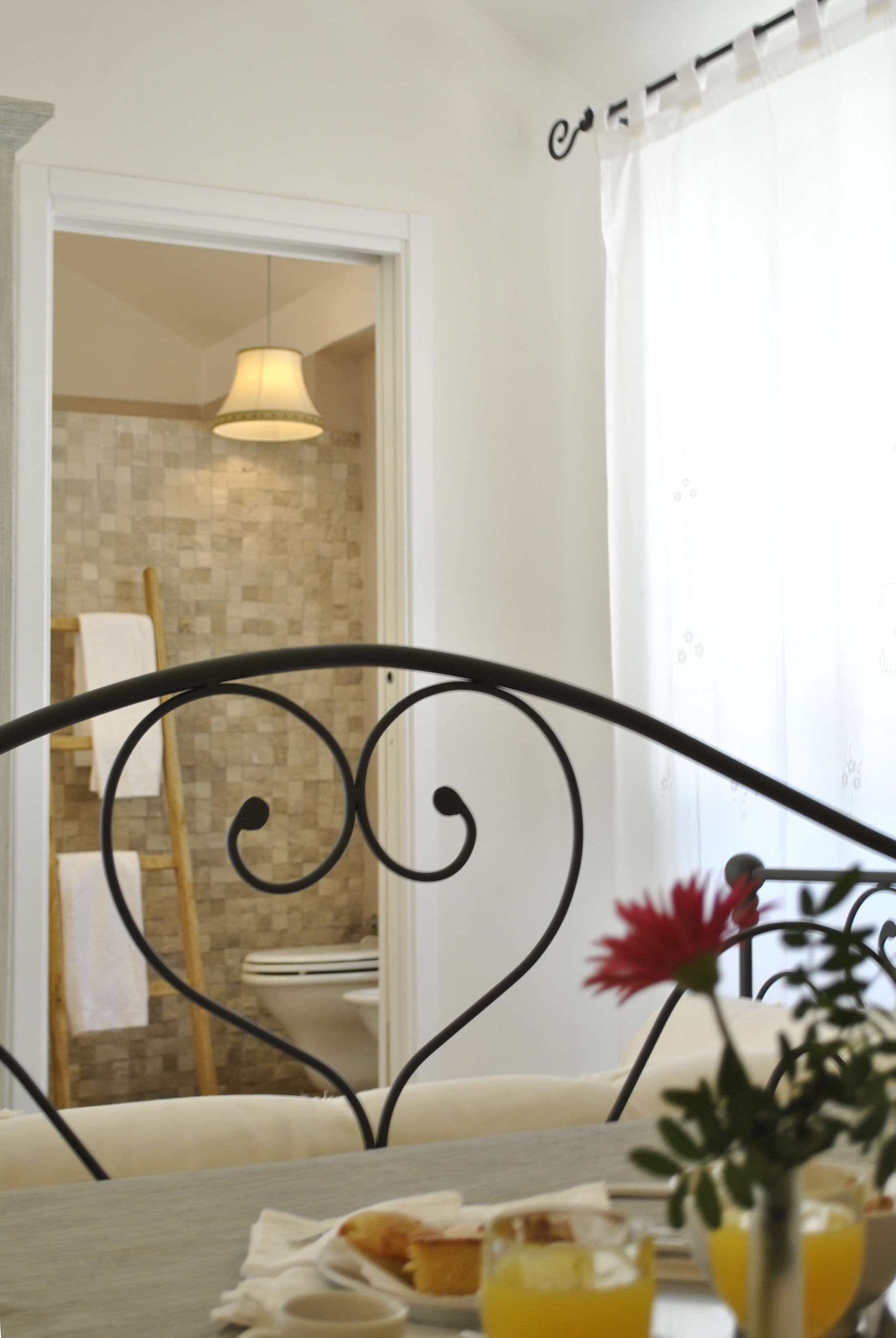 Uma área de refeições com uma mesa posta para o café da manhã, um lustre pendurado e vista para um banheiro com parede revestida de azulejos e um porta-toalhas.