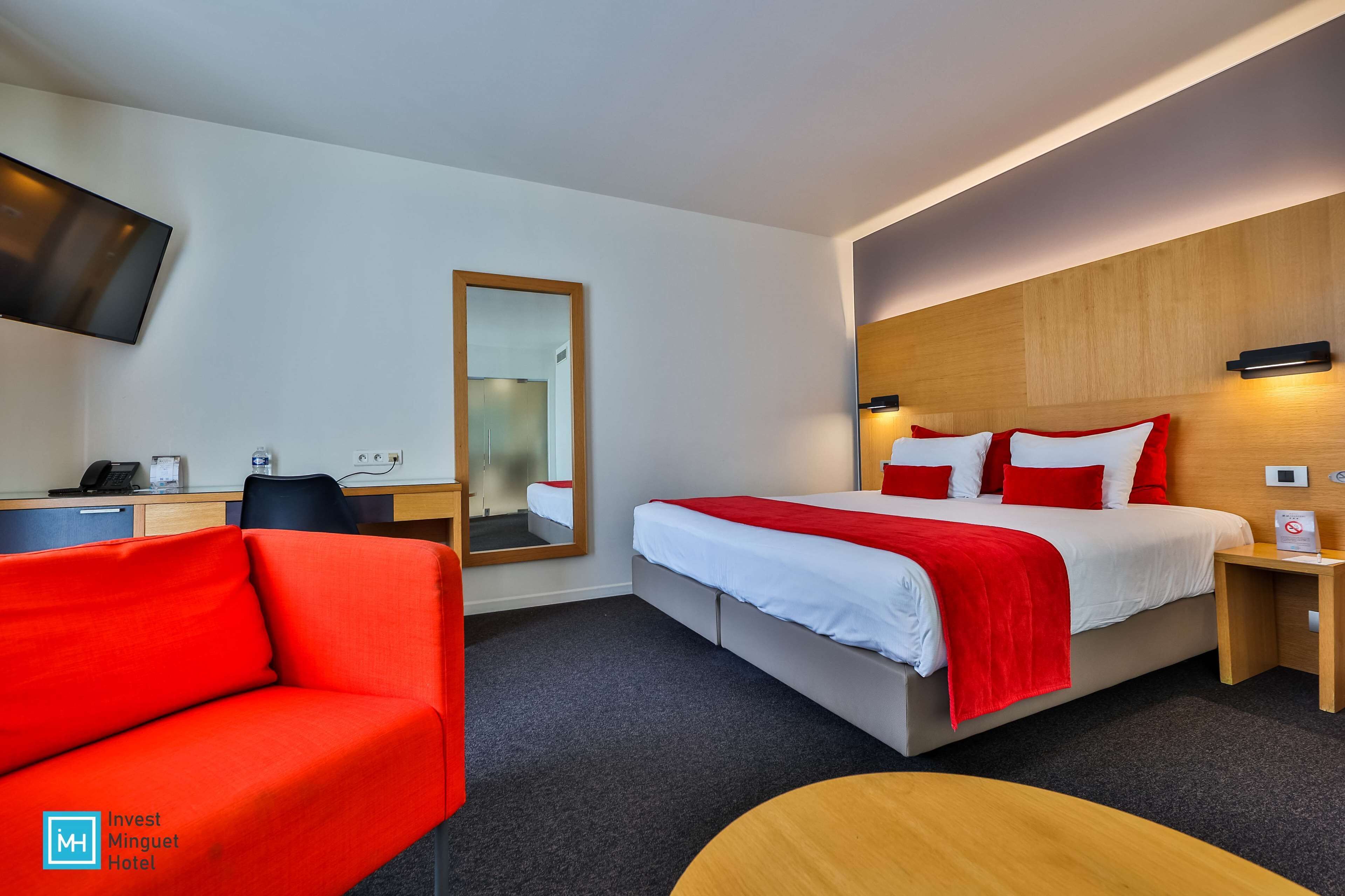Quarto de hotel com cama, escrivaninha, cadeira, televisão e espelho.