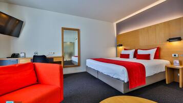 Quarto de hotel com cama, escrivaninha, cadeira, televisão e espelho.