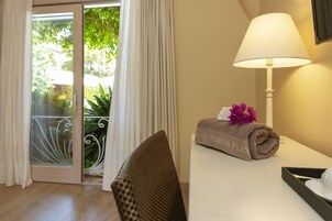 Quarto de hotel com porta de correr de vidro que dá para uma varanda, uma escrivaninha branca com abajur, uma cadeira e uma toalha com a inscrição 'HOTEL SIMPUS PLAY'.