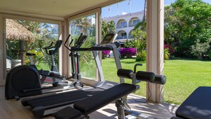 Uma academia com aparelhos de cardio, bancos de musculação e vista para um jardim exuberante e um edifício com bandeiras.