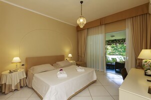 Quarto de hotel com uma cama grande, duas mesas de cabeceira com luminárias, uma escrivaninha com cadeira e vista para o exterior por uma porta de vidro.