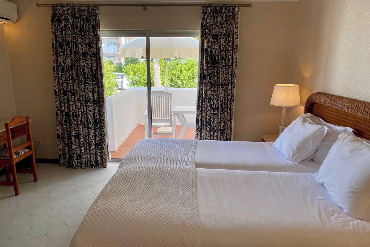 Quarto de hotel com duas camas, uma cadeira, uma mesa e vista para o exterior.