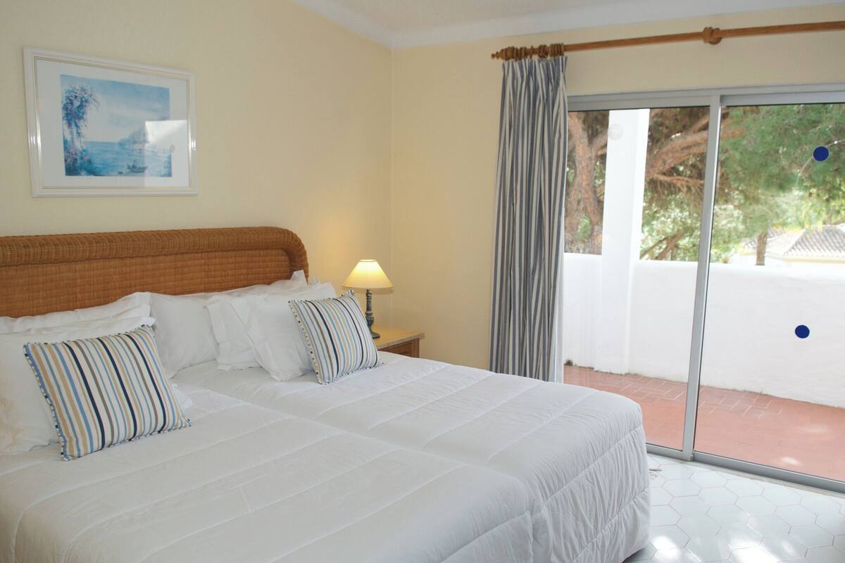 Quarto de hotel com uma cama grande, uma mesa de cabeceira, uma luminária, uma janela com cortinas e um quadro na parede.