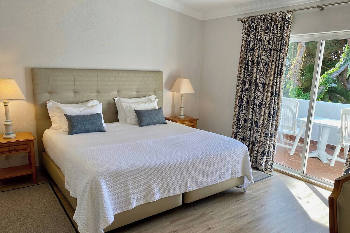 Quarto de hotel com uma cama grande, duas mesas de cabeceira com luminárias, uma janela com cortinas estampadas e vista para um pátio externo.