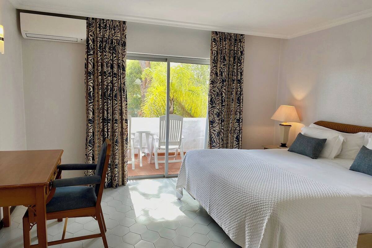 Quarto de hotel com cama, escrivaninha e cadeira. Há uma janela com cortinas, e vista para o verde lá fora.