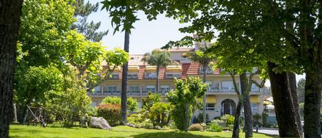 Um hotel com fachada amarela, várias varandas e um jardim bem cuidado com árvores e arbustos.