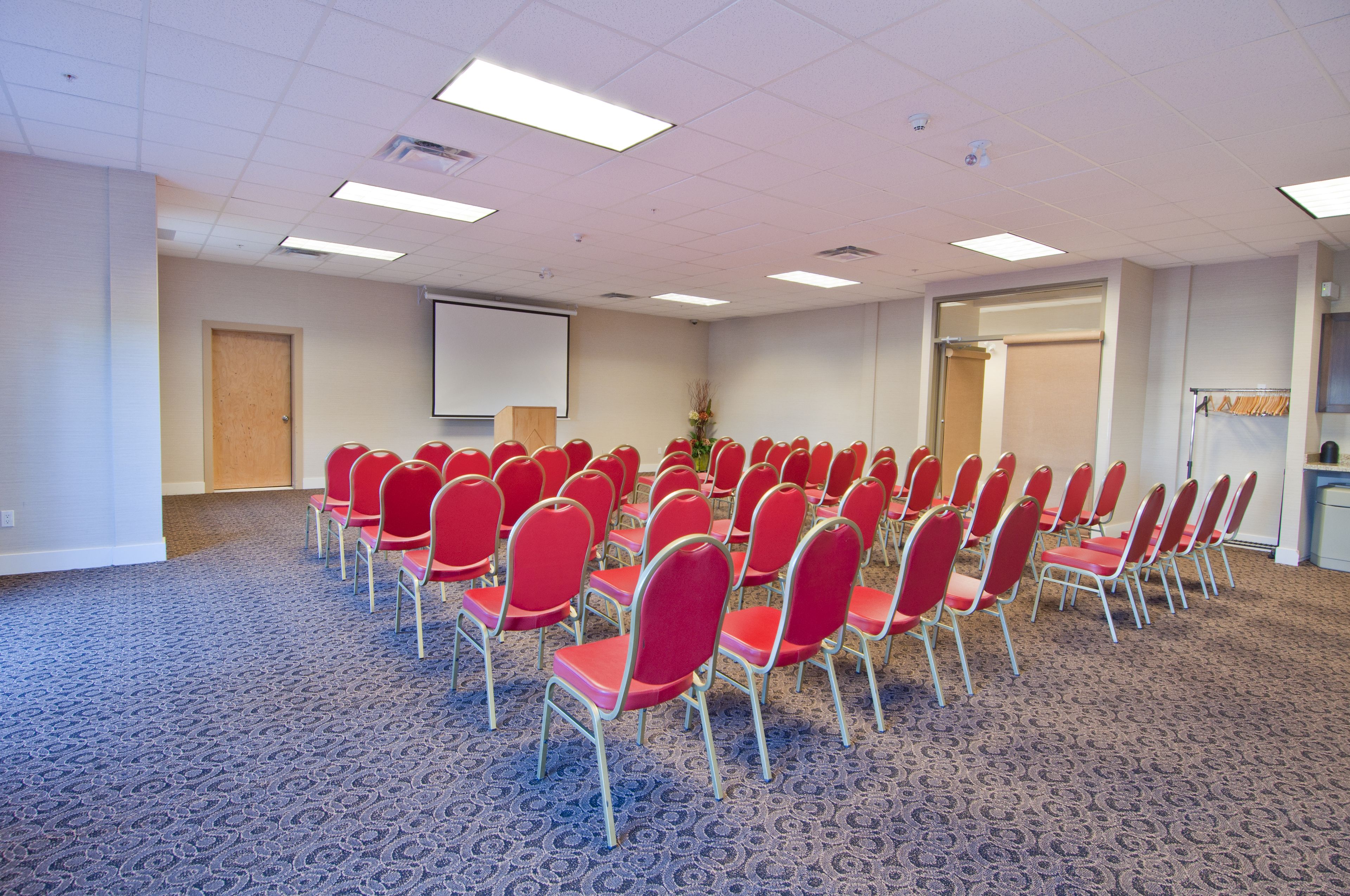 Uma sala de conferências com fileiras de cadeiras vermelhas, uma tela de projetor e piso carpeteado.
