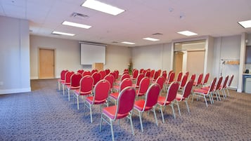 Uma sala de conferências com fileiras de cadeiras vermelhas, uma tela de projetor e piso carpeteado.