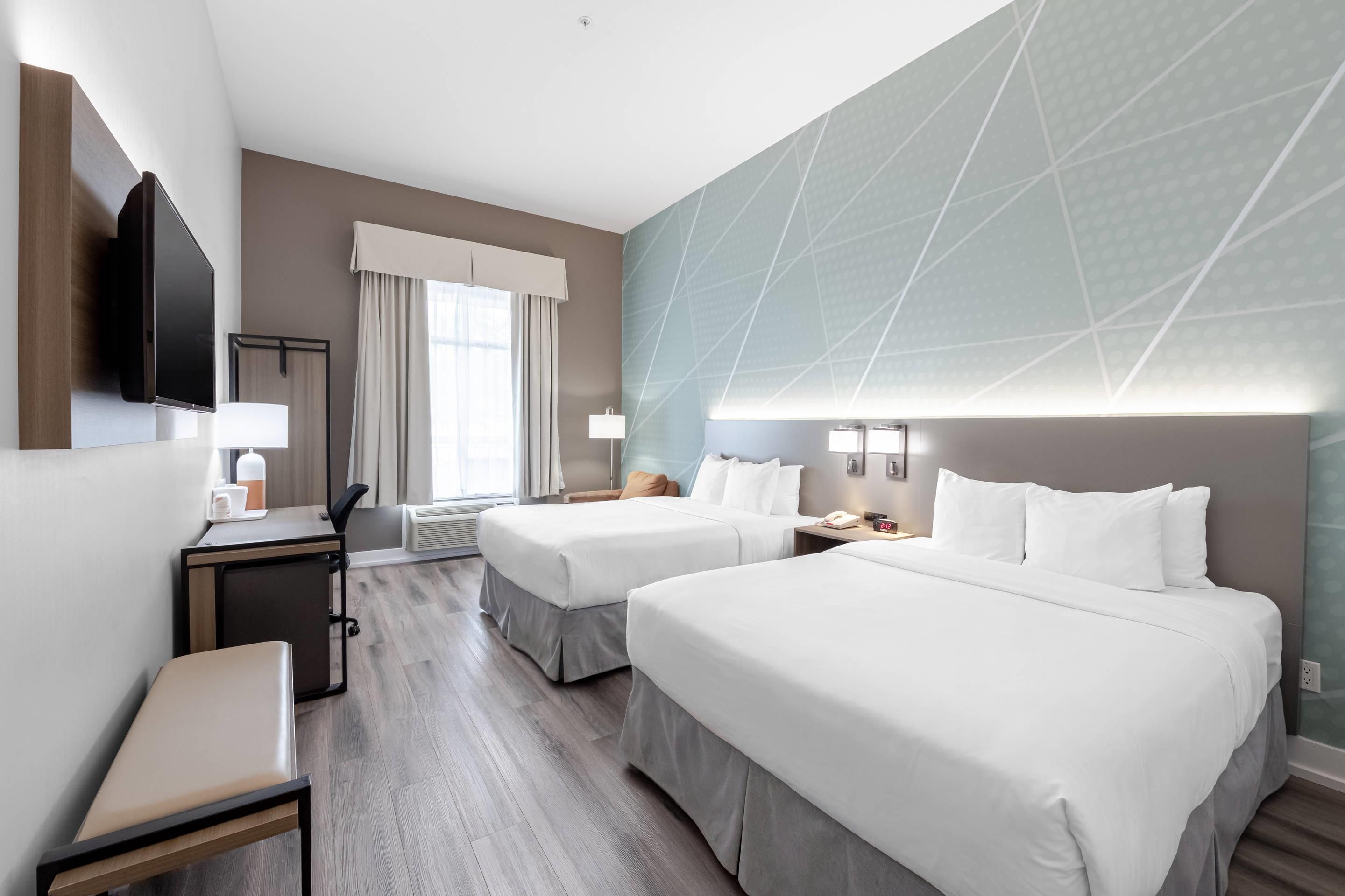 Quarto de hotel com duas camas, uma mesa de trabalho com cadeira, televisão e janela com cortinas.