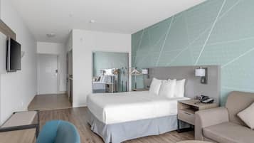 Quarto de hotel moderno com uma cama grande, escrivaninha, cadeira e sofá. Há um espelho, uma TV e um telefone na parede.