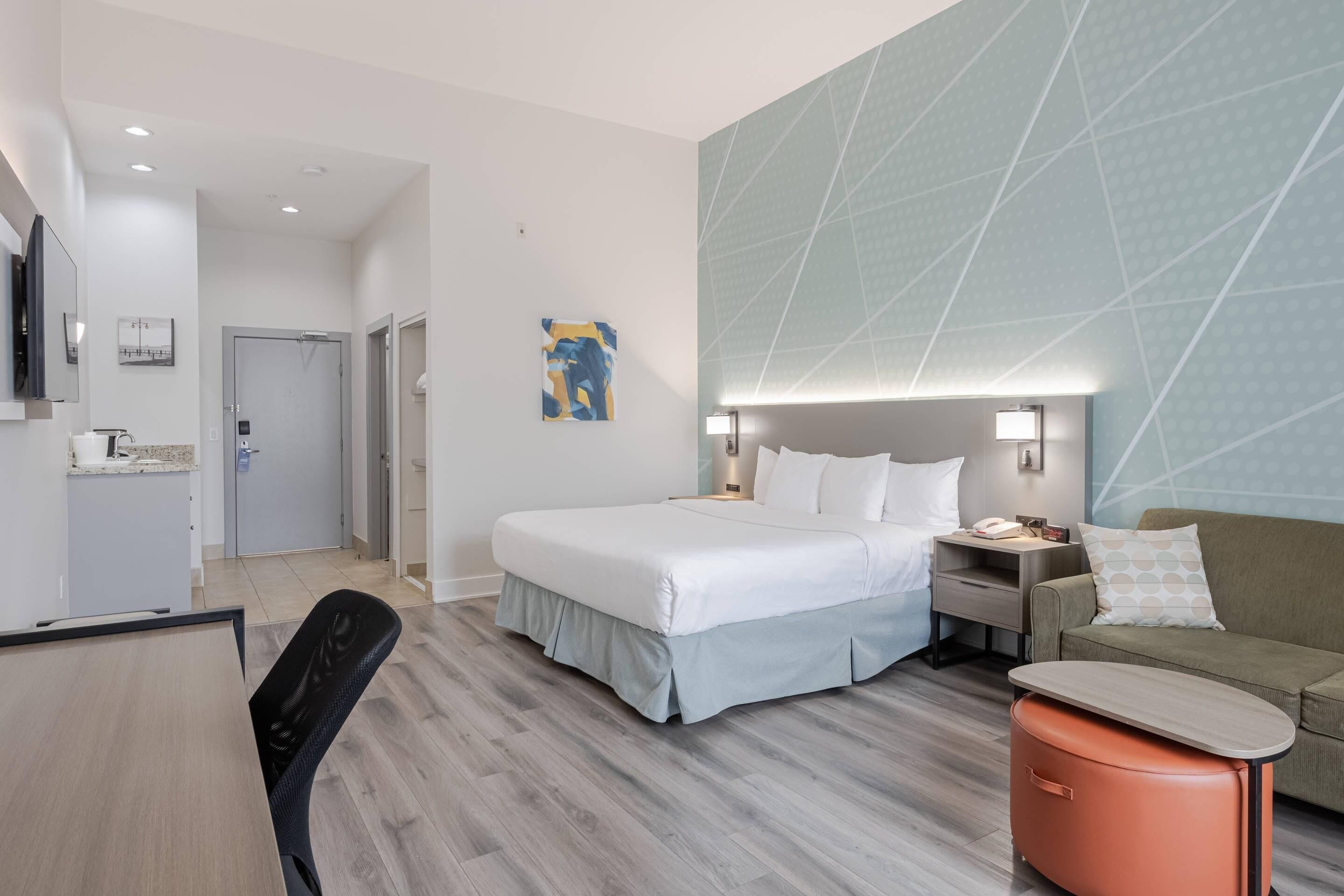 Quarto de hotel com cama, mesa de cabeceira, abajur e um pequeno banquinho laranja.