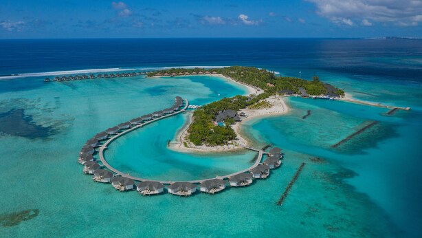 Vista aérea de uma ilha resort com bangalôs sobre a água, ligados por uma passarela sinuosa, rodeada por águas azul-turquesa e um céu claro.
