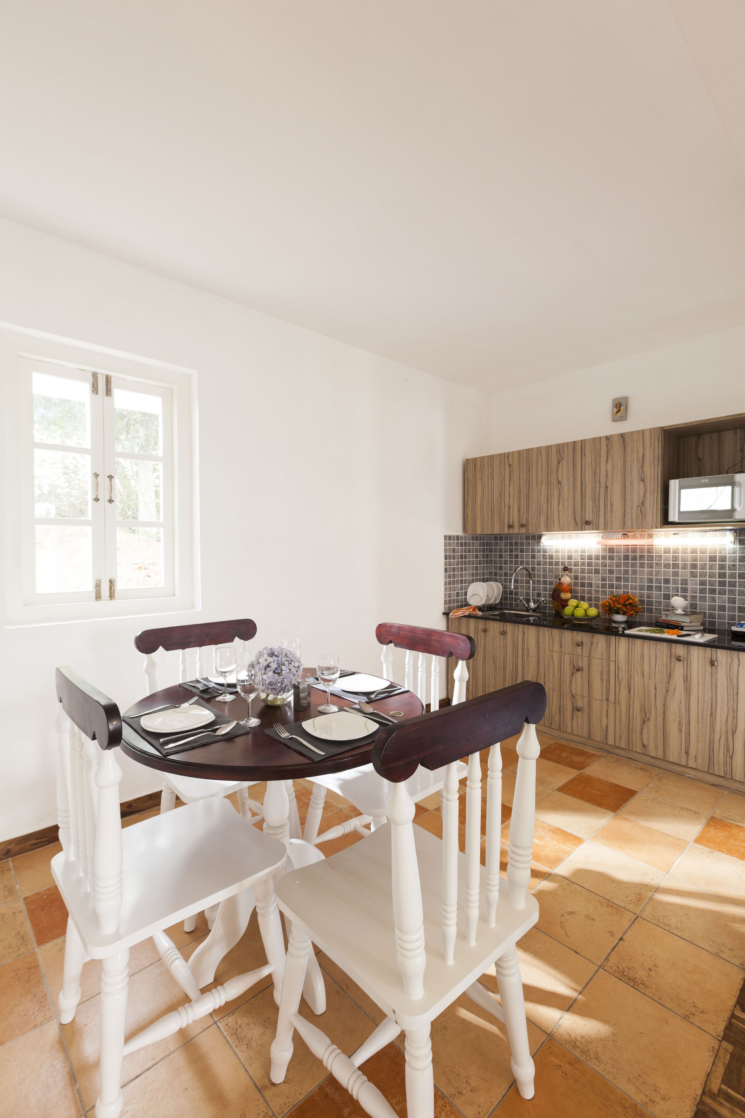 Uma sala de jantar com uma mesa redonda e cadeiras brancas, uma cozinha com armários de madeira e painel de azulejos, e uma janela com persianas brancas.