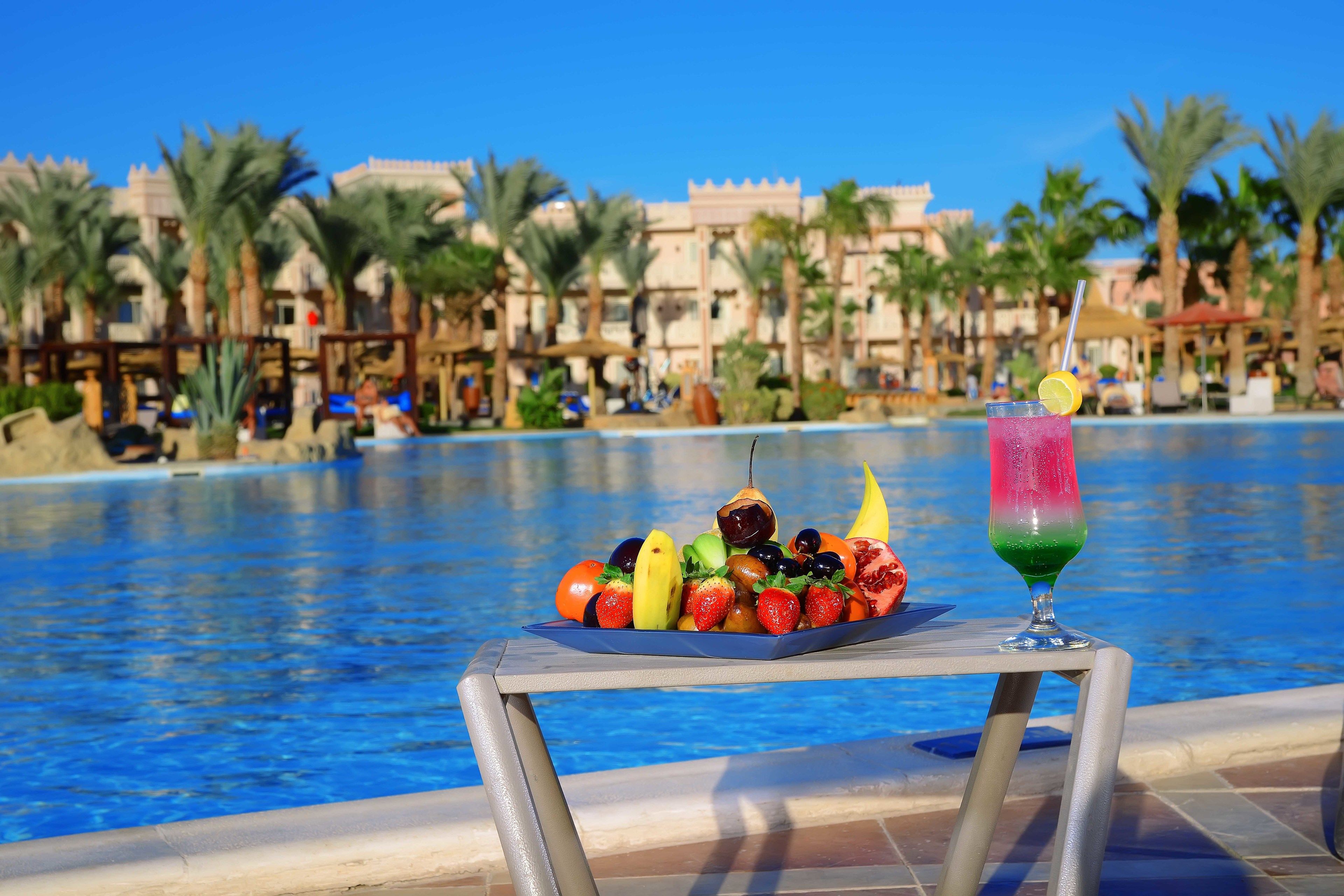 Mesa à beira da piscina com uma travessa de frutas e uma bebida colorida, rodeada por palmeiras e piscina.