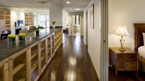 Corredor moderno de hotel com piso de madeira, área de cozinha com bancada e armários, e quarto com mesa de cabeceira e luminária.