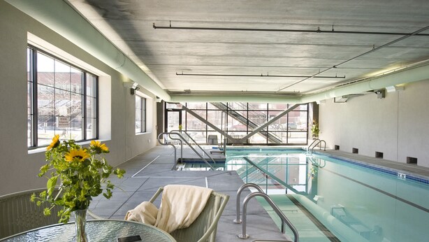 Uma área interna de piscina moderna com paredes de vidro transparente, uma cadeira de descanso, uma mesa com um vaso de flores e uma escadaria que leva a um andar superior.
